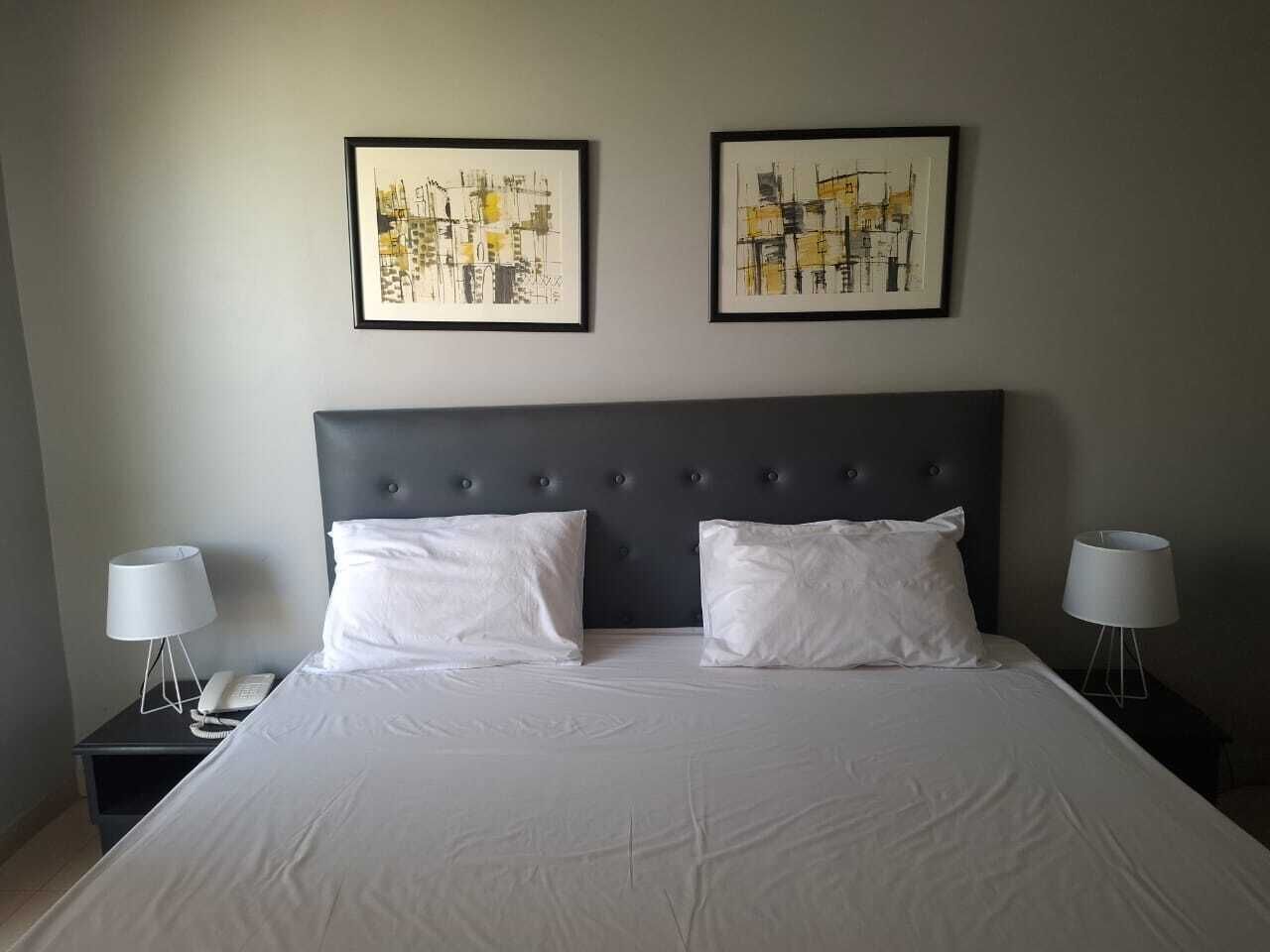 Quarto de hotel com uma cama grande, duas almofadas brancas, cabeceira escura e duas obras de arte emolduradas na parede.