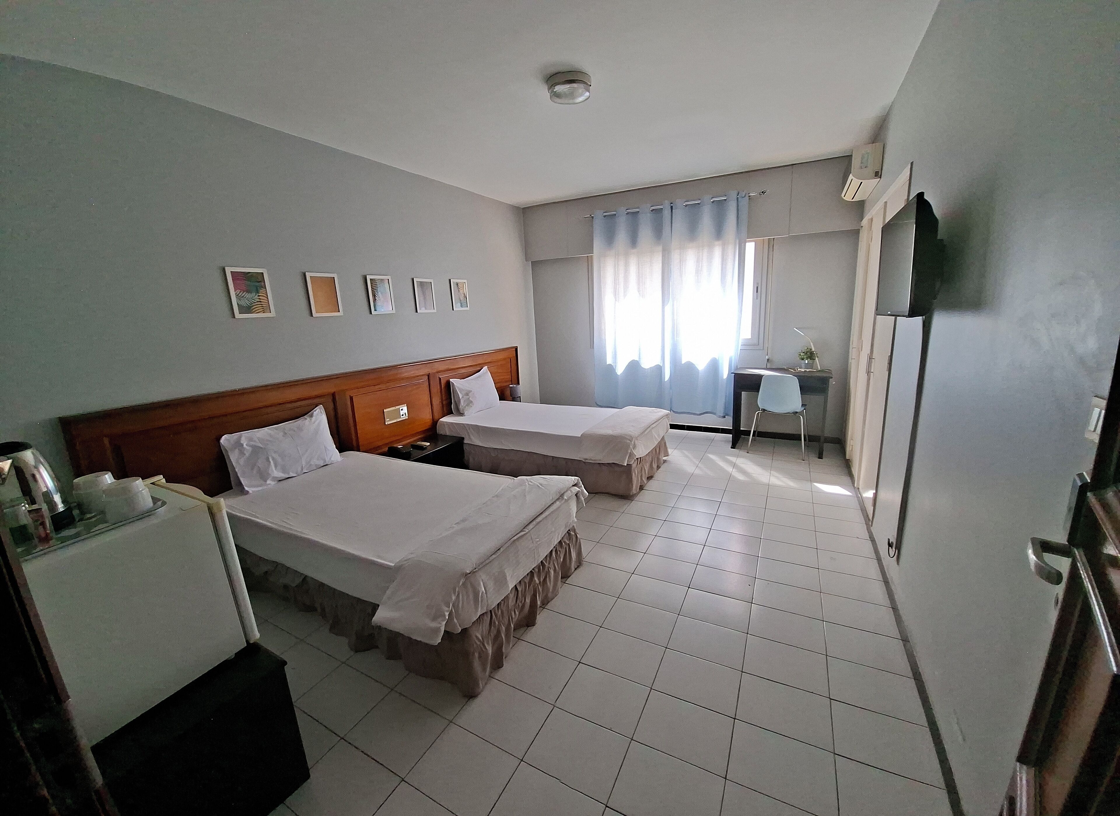 Quarto de hotel com duas camas, uma escrivaninha, uma cadeira, um refrigerador e uma janela com cortinas.
