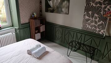 Quarto com cama, prateleira de madeira com caixas estampadas, radiador e janela com vista.
