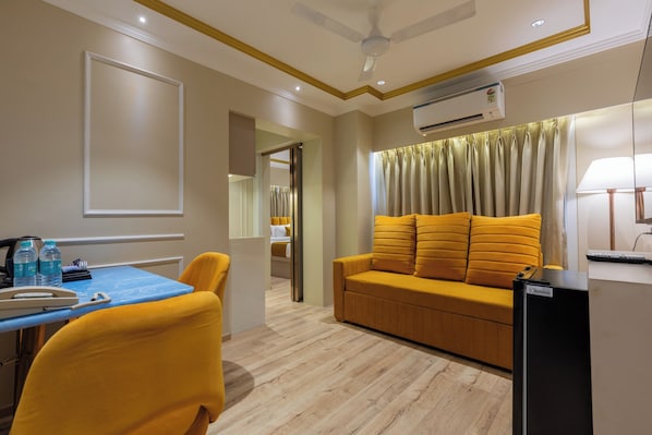 Quarto de hotel com piso de madeira, um sofá amarelo, uma mesa azul com telefone e um ar condicionado.