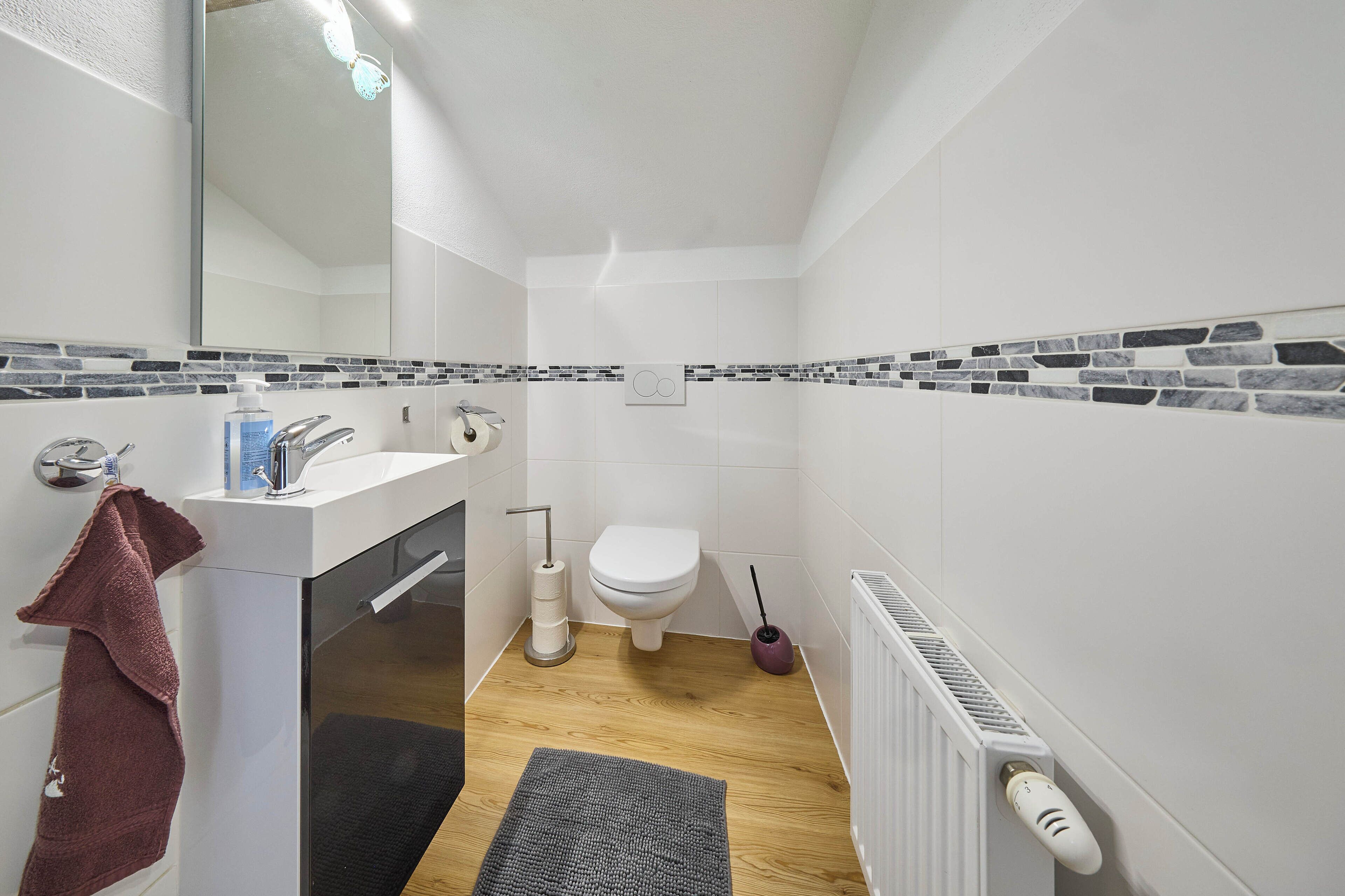 Casa de banho moderna com lavatório branco, espelho e sanita. Tem um porta-toalhas com toalha, escova de sanita e radiador.