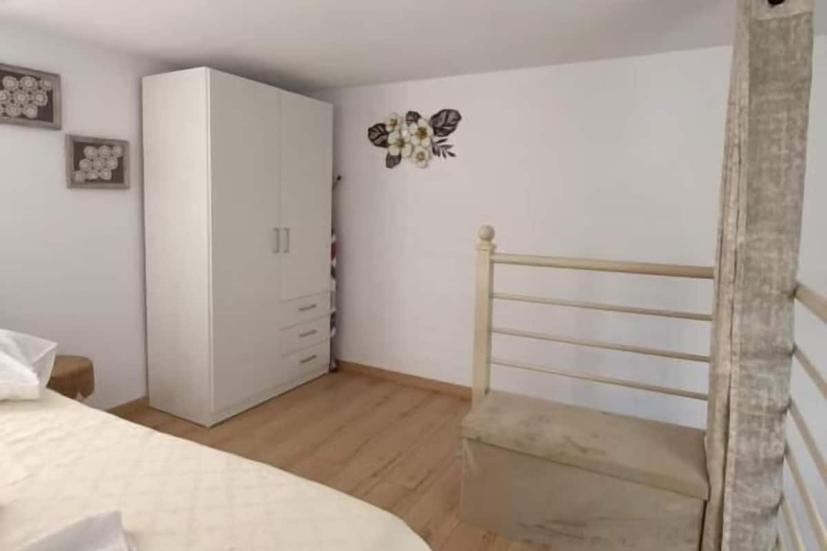 Um quarto com uma cama, um guarda-roupa, um banquinho pequeno e uma cadeira de madeira.