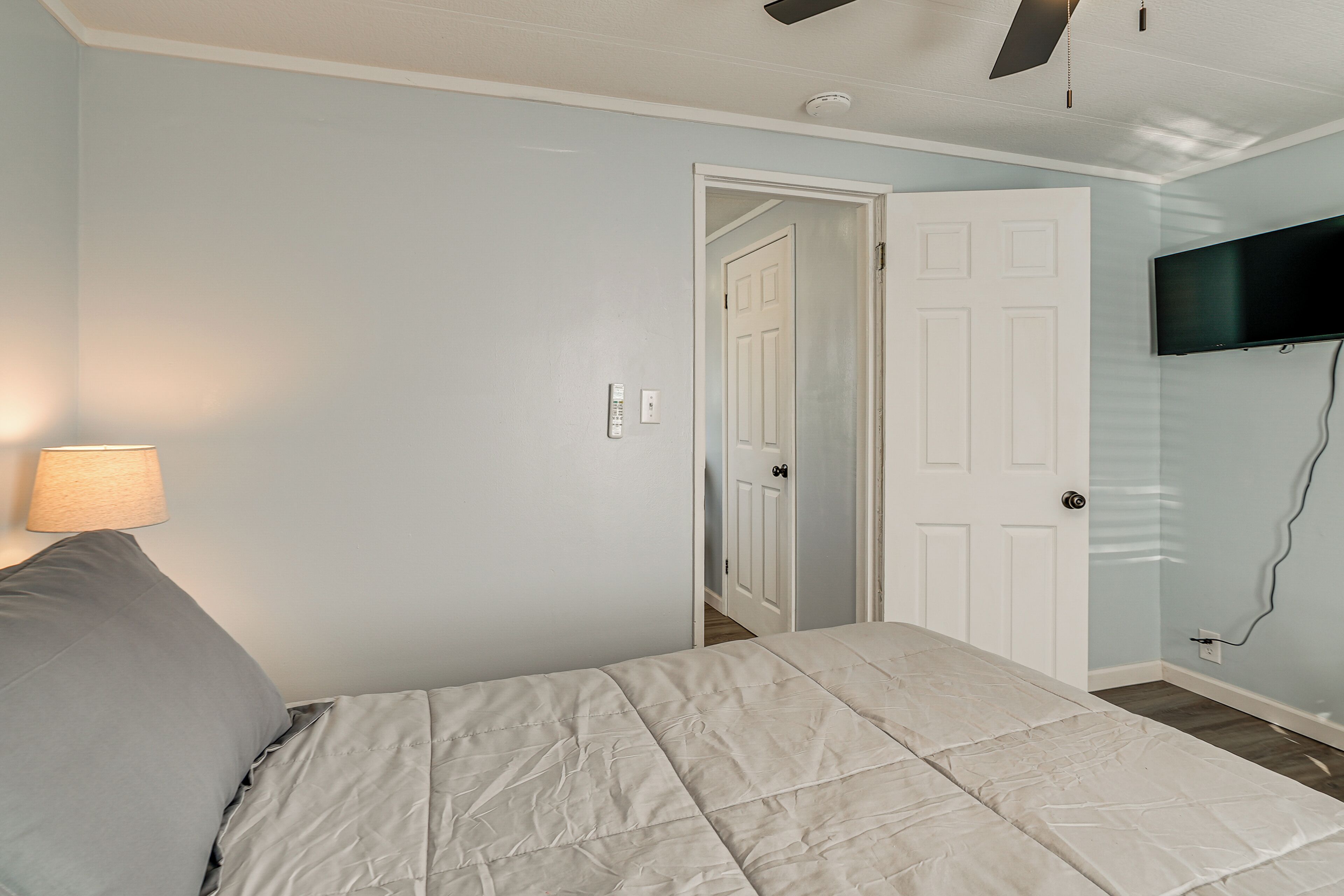 Quarto com cama, luminária, TV fixada na parede e porta que dá para outro cômodo.