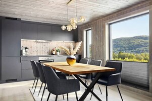 Cozinha moderna com armários escuros, uma mesa de jantar de madeira e cadeiras. Há uma janela grande com vista para uma área arborizada.
