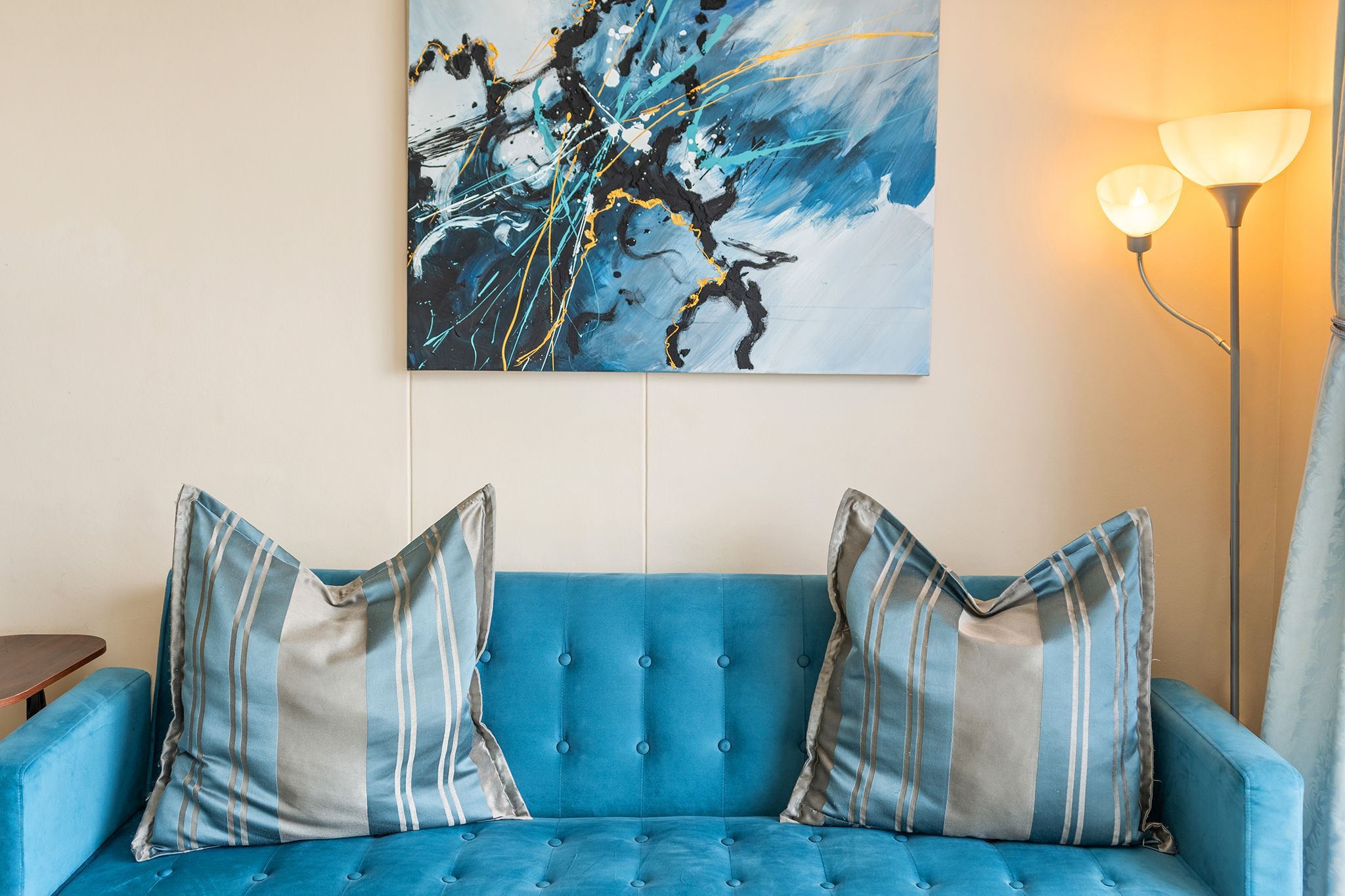 Um sofá azul com almofadas listradas, uma pintura abstrata moderna e um abajur.