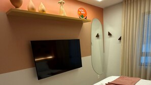 Quarto de hotel com cama, televisão de tela plana, espelho, prateleira com objetos decorativos e janela com cortinas.