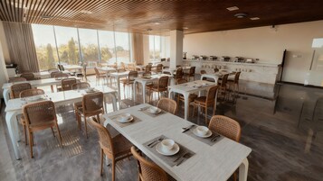 Restaurante moderno com ambiente amplo, caracterizado por pé direito alto, grandes janelas e diferentes opções de assentos, incluindo mesas e cadeiras.