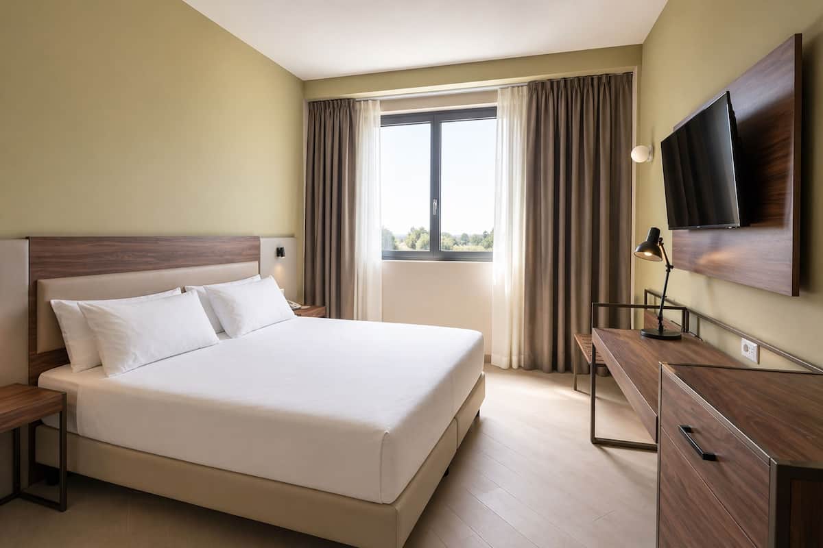 Quarto de hotel com cama, mesa de cabeceira, escrivaninha, televisão e janela grande com cortinas.