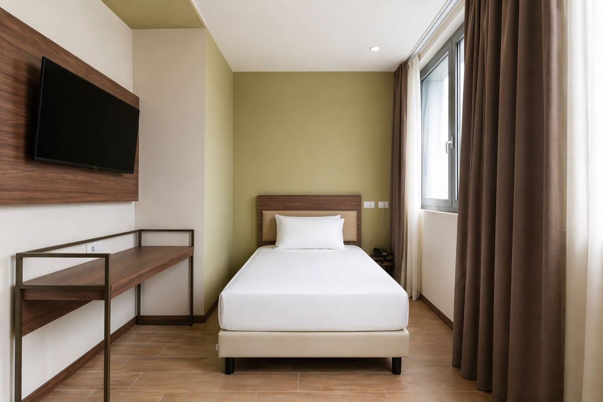 Quarto de hotel com uma cama, televisão na parede, escrivaninha e janela com cortinas.