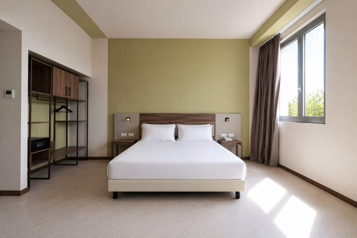 Quarto de hotel moderno com uma cama grande, um guarda-roupa, uma mesa de cabeceira e uma janela com cortinas.
