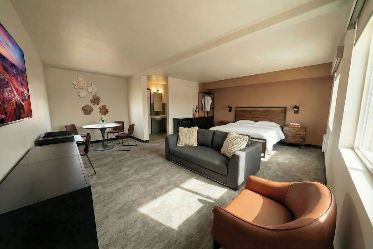 Quarto de hotel com cama, sofá, escrivaninha e cadeira. Uma janela permite entrada de luz natural.