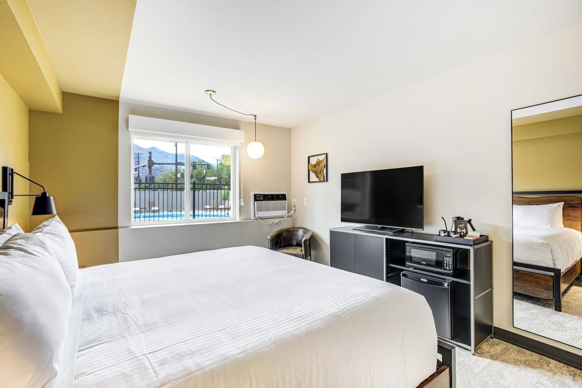 Quarto de hotel com cama, televisão, escrivaninha com micro-ondas e vista para uma piscina através de uma janela.