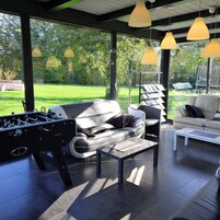Sala de estar moderna com mesa de futebol, dois sofás e uma mesa de centro. Grandes janelas oferecem vista para um gramado verde e árvores.