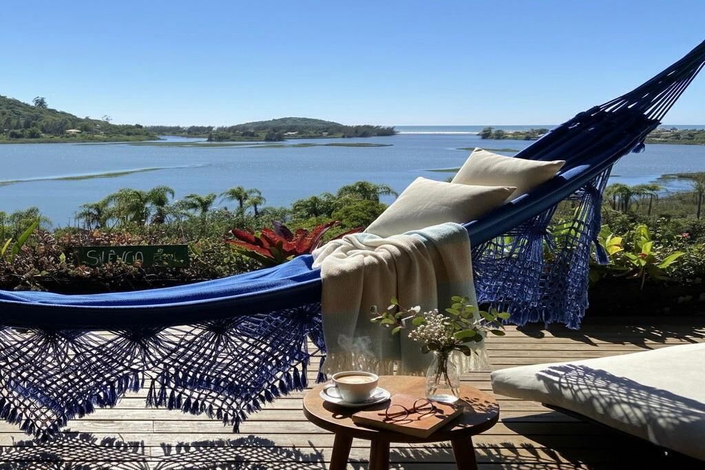 Uma cadeira rede com almofadas, vista para um lago, um deck de madeira e uma mesa com uma xícara e flores.