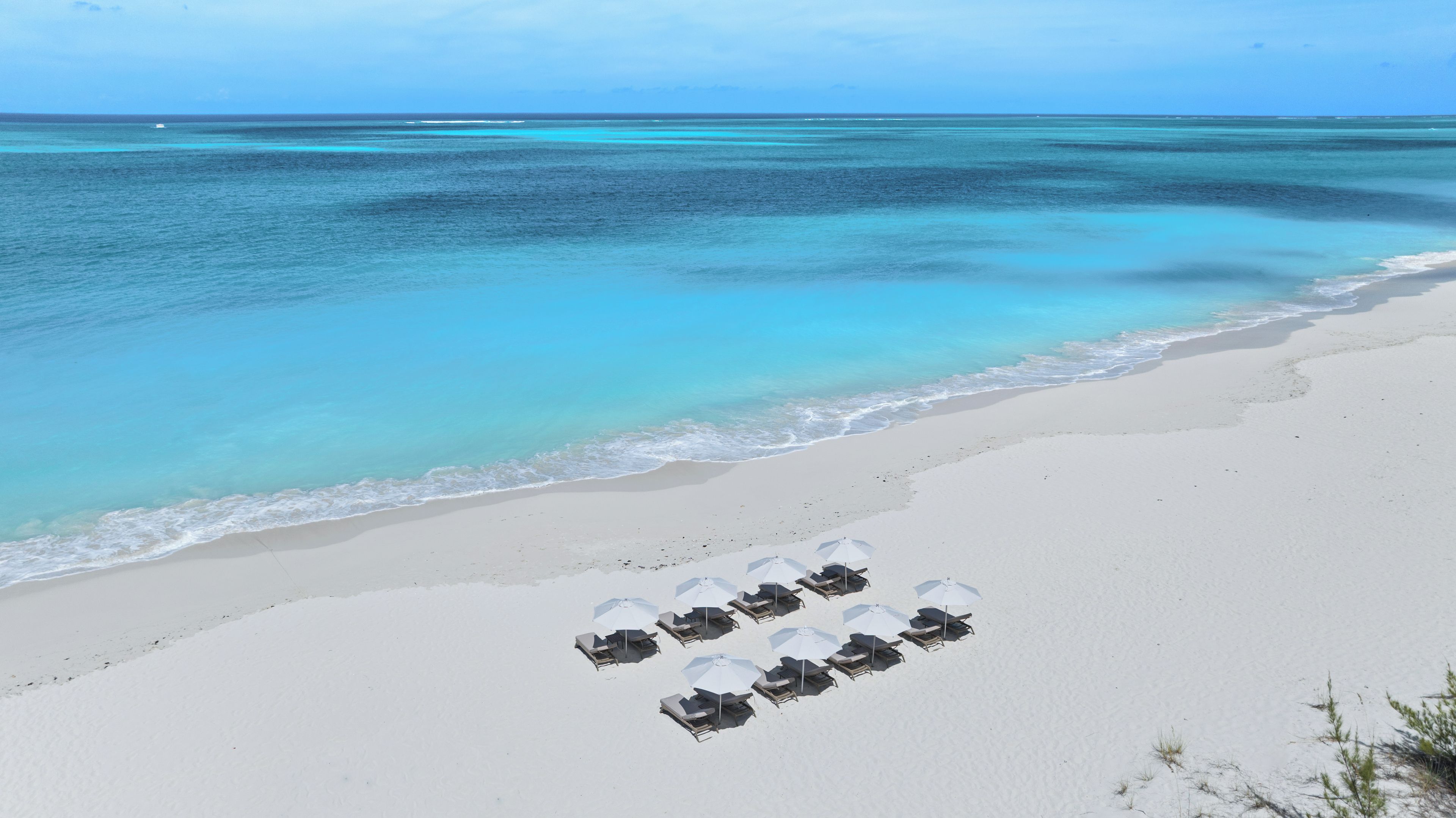 Praia com areia branca e águas turquesa cristalinas. Várias cadeiras de praia e guarda-sóis estão dispostos na areia.