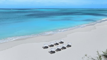 Praia com areia branca e águas turquesa cristalinas. Várias cadeiras de praia e guarda-sóis estão dispostos na areia.