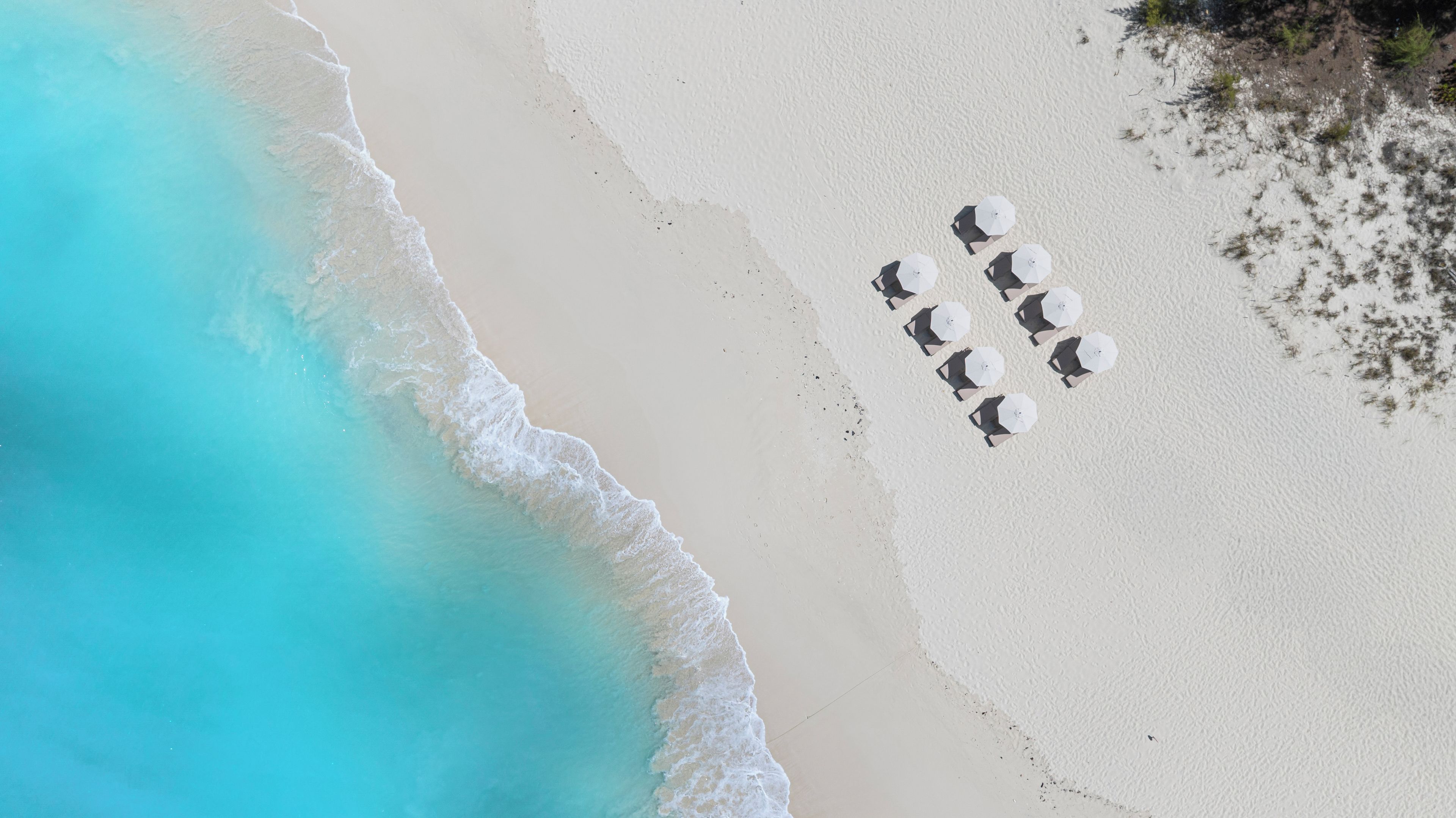 Vista aérea de uma praia com areia branca e água turquesa. Vários guarda-sóis brancos estão dispostos em linha na areia.