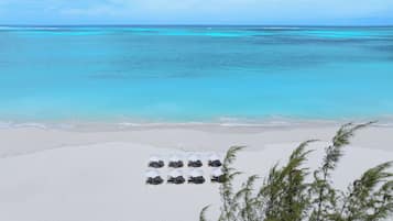 Uma praia com areia branca e água turquesa. Fileiras de guarda-sóis e cadeiras brancas estão dispostas.