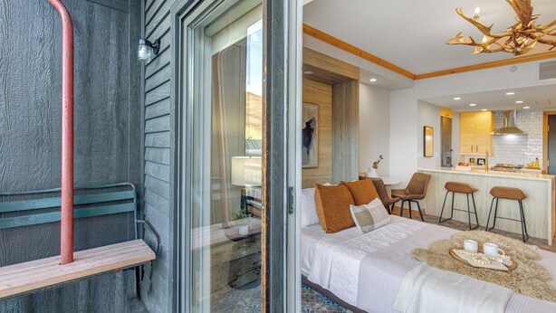 Quarto de hotel moderno com porta de vidro, cama com roupa de cama branca, um banco de madeira e uma cozinha com banquetas.