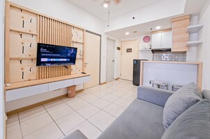 Uma sala moderna com televisão de tela plana fixada em uma parede de madeira, um sofá cinza e uma área de cozinha com armários brancos e painel de azulejos.
