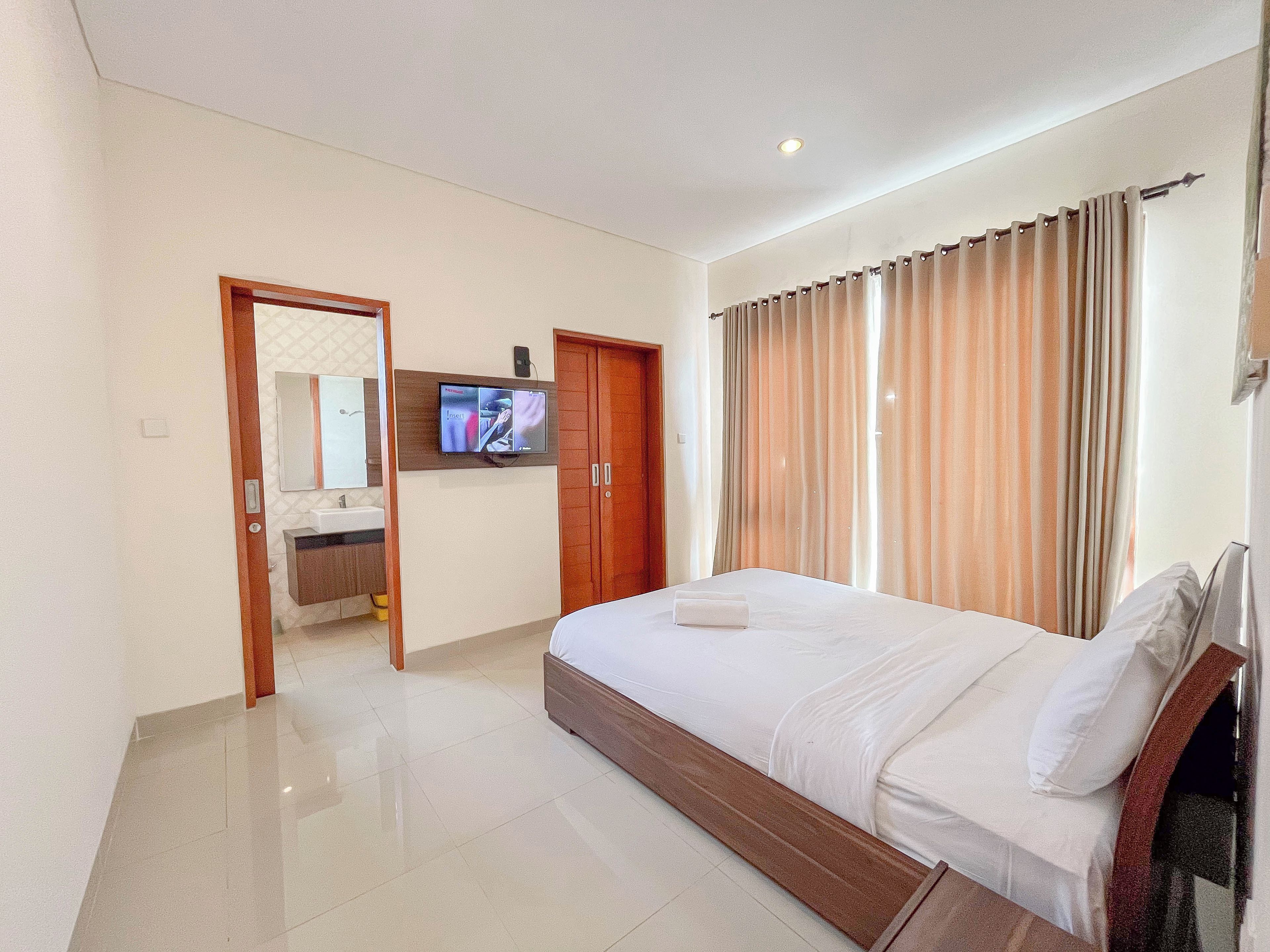 Quarto de hotel com cama, televisão presa na parede, banheiro com pia e porta que dá para outro cômodo.
