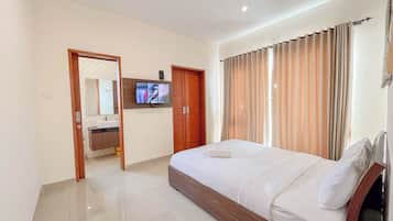 Quarto de hotel com cama, televisão presa na parede, banheiro com pia e porta que dá para outro cômodo.