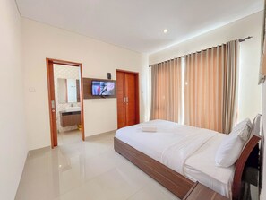 Quarto de hotel com cama, televisão presa na parede, banheiro com pia e porta que dá para outro cômodo.