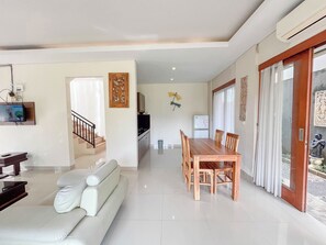 Uma sala de estar moderna com sofá, mesa de jantar e cadeiras. Há uma escada que leva a outro nível, uma área de cozinha e um espaço de refeições com vista para o exterior.