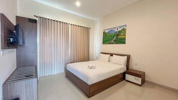 Quarto de hotel com cama, televisão na parede, um quadro e uma mesinha de cabeceira.