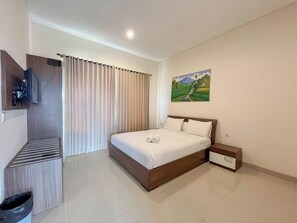 Quarto de hotel com cama, televisão na parede, um quadro e uma mesinha de cabeceira.