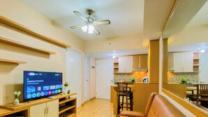 Sala de estar com televisão, móveis de madeira, ventilador de teto e área de cozinha ao fundo.