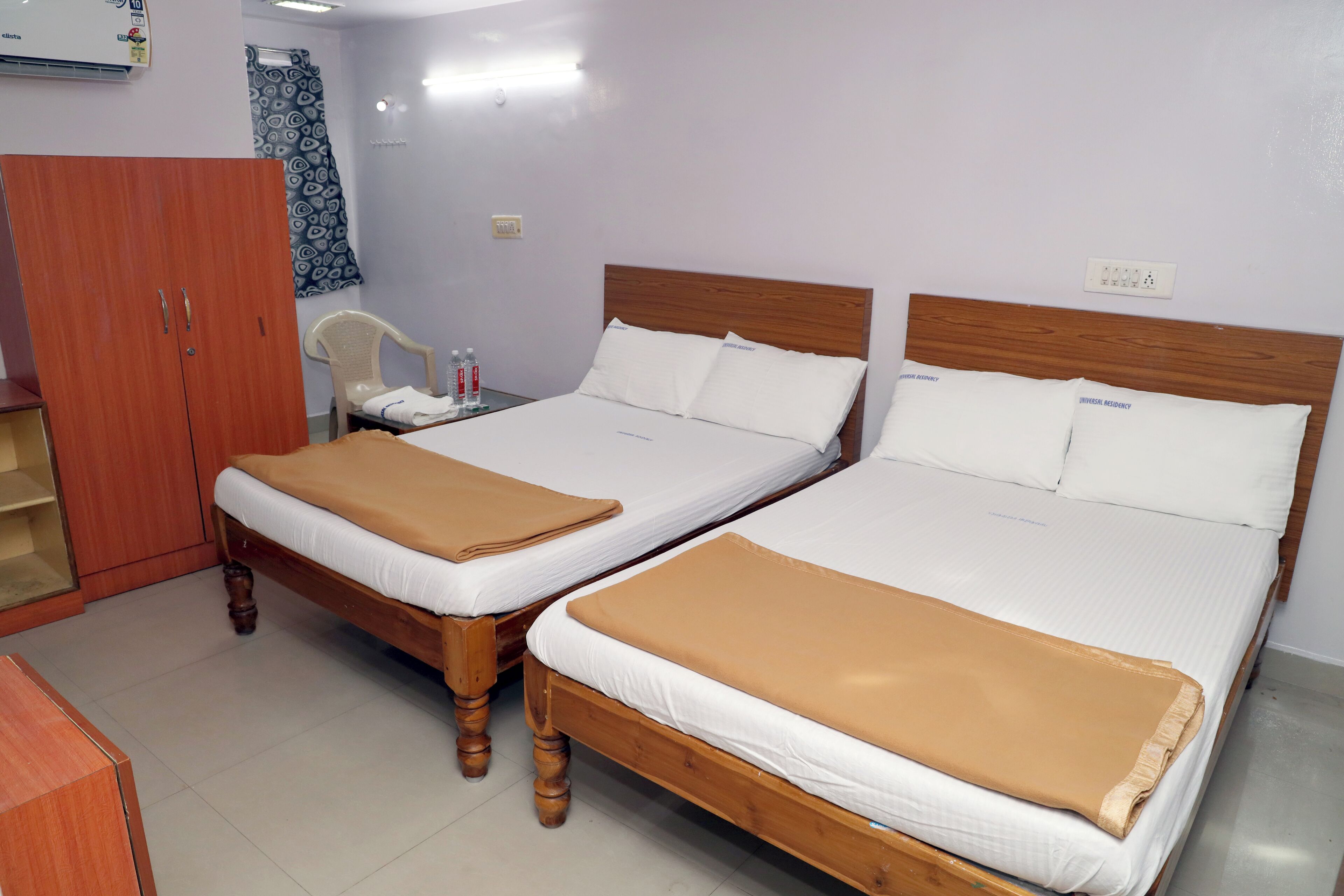 Quarto de hotel com duas camas de madeira, um guarda-roupa, uma cadeira e uma mesa de cabeceira com garrafas.