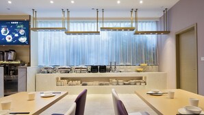 Uma sala de jantar moderna com mesas preparadas para refeições, área de cozinha com equipamentos de serviço de alimentos e uma grande janela com cortinas.
