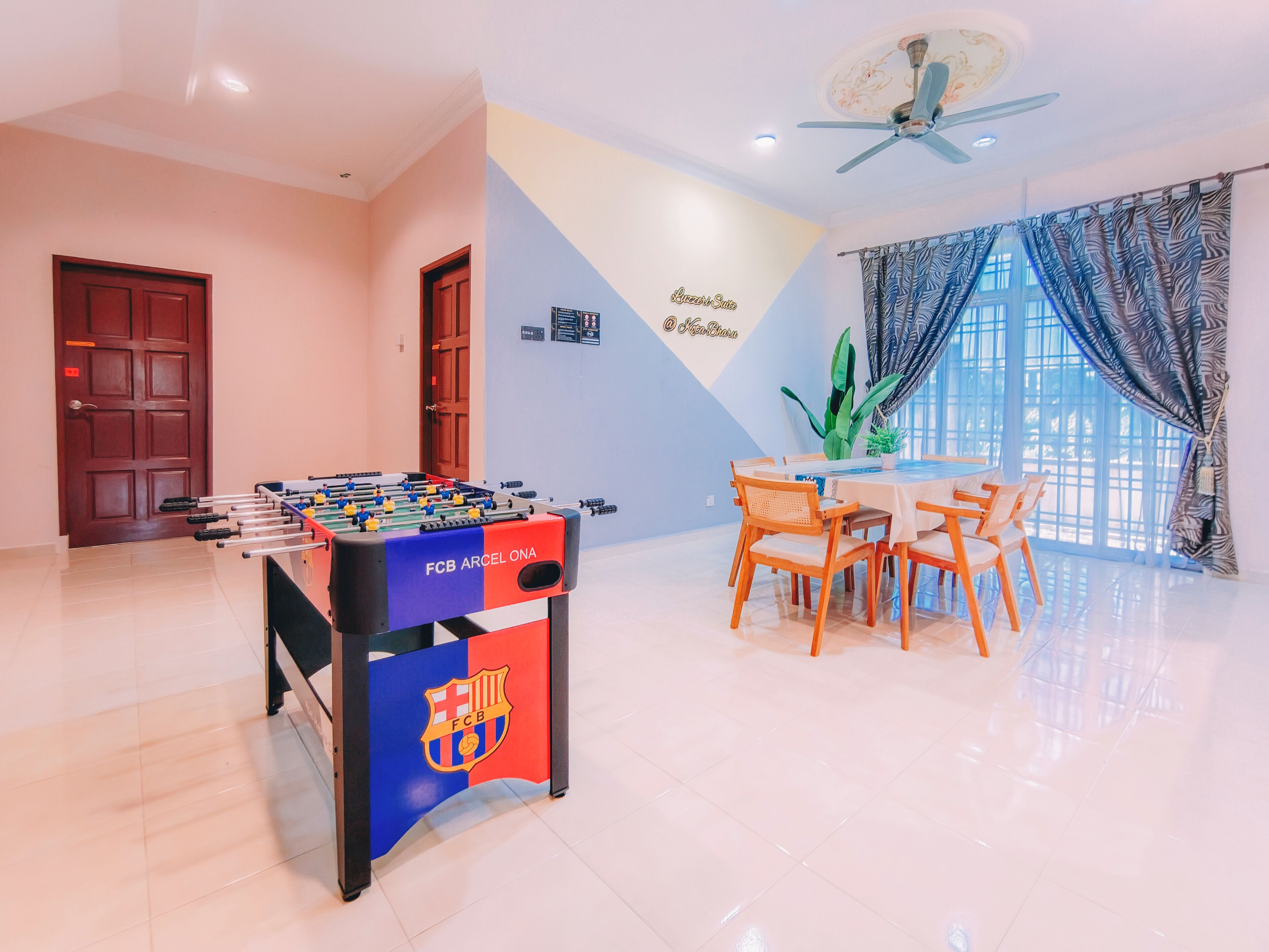 Quarto com mesa de pebolim, mesa de jantar com cadeiras, ventilador de teto e janela com cortinas.