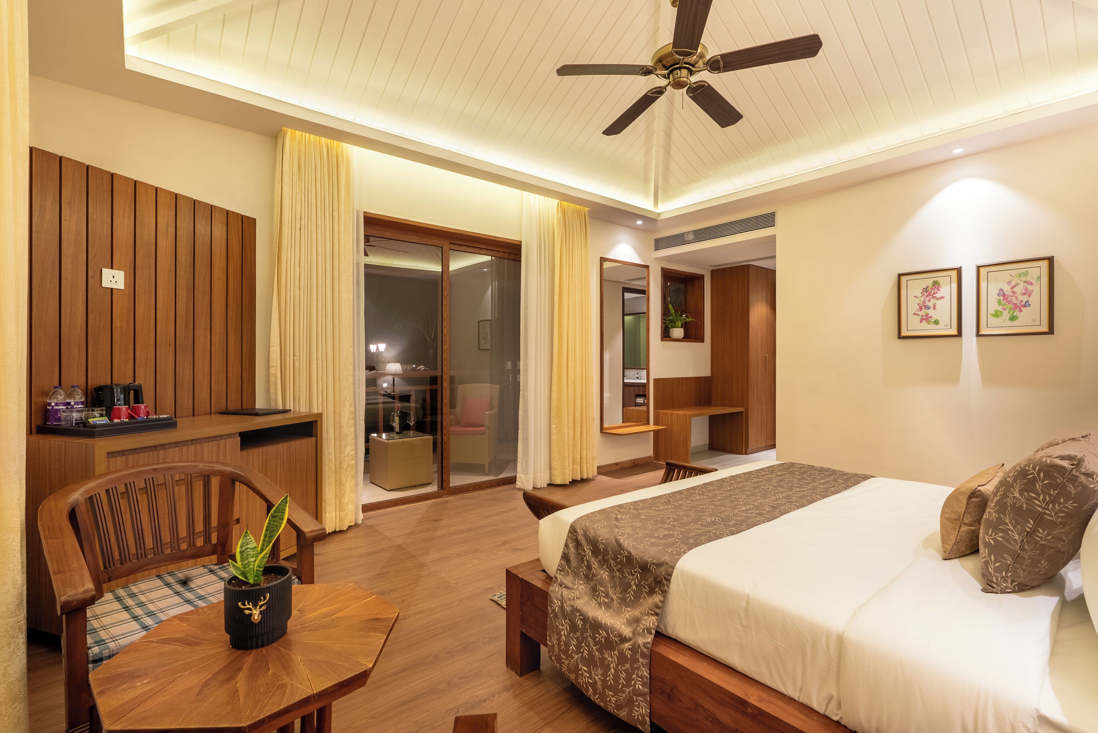 Quarto de hotel com cama, escrivaninha, cadeira, planta em vaso e ventoinha no teto.
