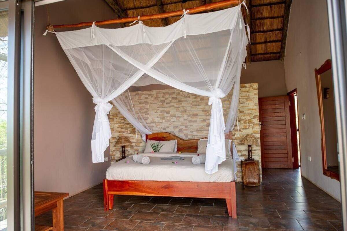 Quarto com cama sob um dossel branco, parede de pedra, porta de madeira e uma pequena mesa.