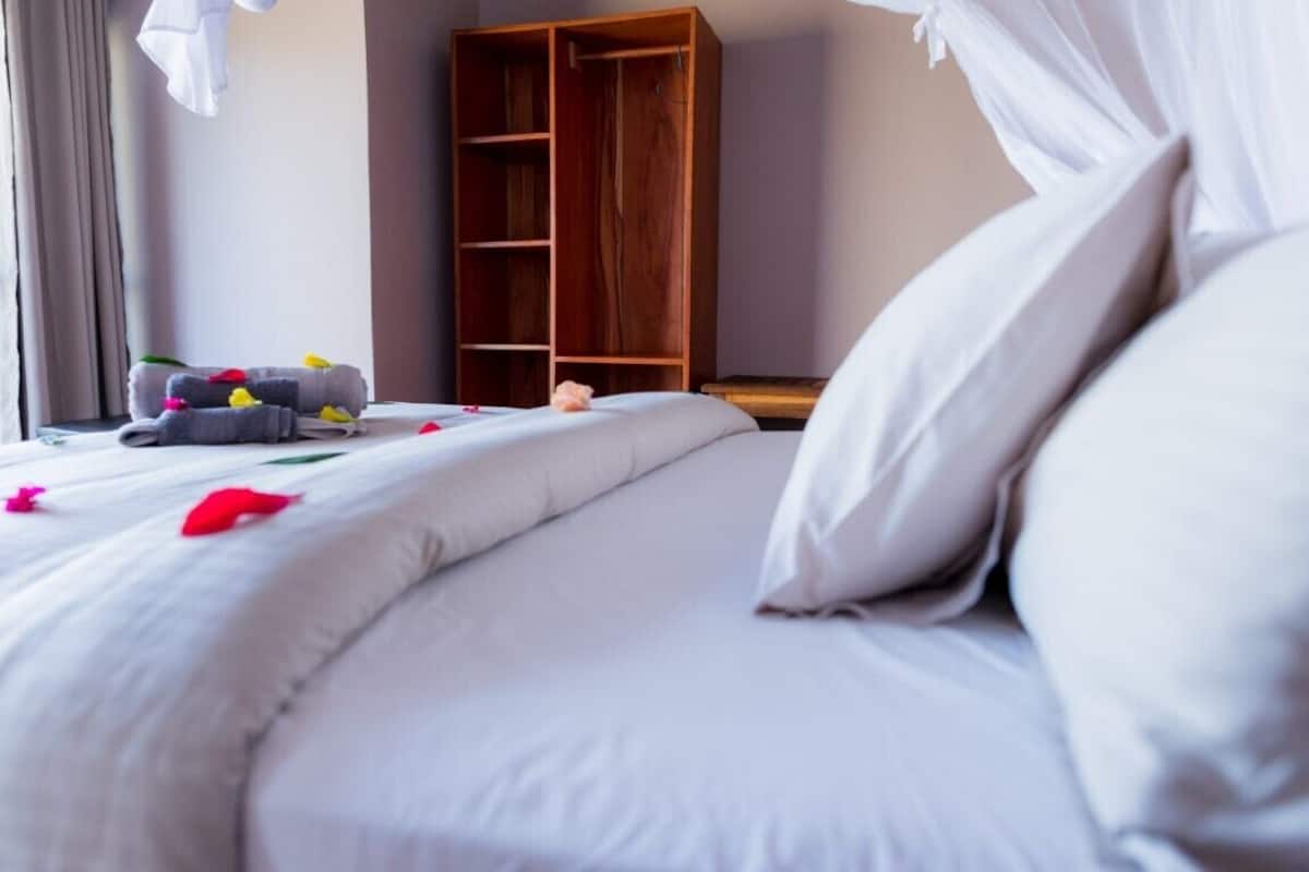 Uma cama bem arrumada com lençóis e almofadas brancas, um guarda-roupa de madeira e uma mesa de cabeceira com abajur.