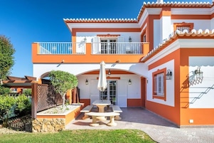 Um edifício de dois andares com um pátio que inclui uma mesa, cadeiras, um espaço de estar coberto e uma parede de pedra.