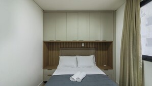 Quarto de hotel com cama, cabeceira de madeira, mesa de cabeceira e janela com cortinas.
