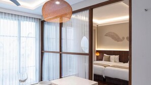 Quarto de hotel com área de refeições, uma grande janela com cortinas, cama com cabeceira e luminária fixada na parede.