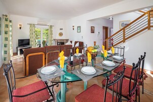Uma sala de jantar com uma mesa comprida posta para seis pessoas, rodeada por cadeiras estofadas em vermelho. Uma televisão está visível ao fundo, e uma escada leva a um andar superior.
