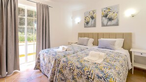 Quarto com uma cama grande, duas mesas de cabeceira, uma janela com cortinas e dois quadros florais na parede.