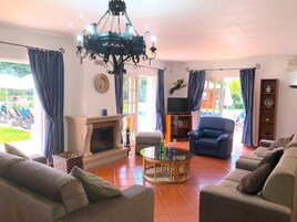 Uma sala de estar com lareira, uma mesa de centro redonda, sofás, uma poltrona azul e um candelabro.