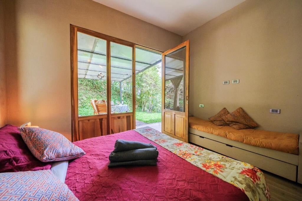 Quarto com cama, um banquinho e porta de correr para área externa.