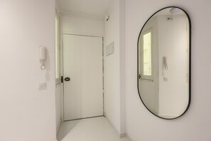 Um corredor com uma porta branca, um espelho oval fixado na parede e um telefone branco também fixado na parede.