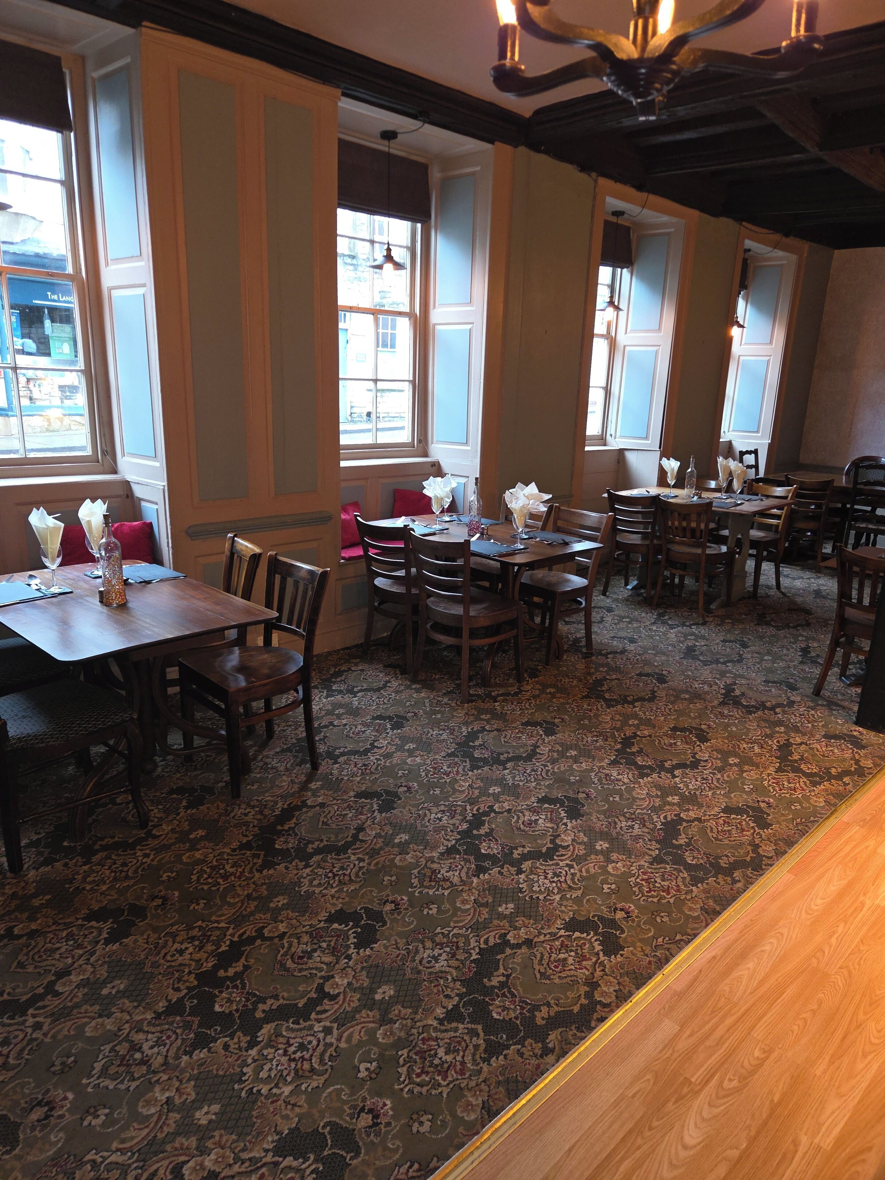 Interior de restaurante com mesas e cadeiras de madeira, janelas amplas e um tapete estampado.