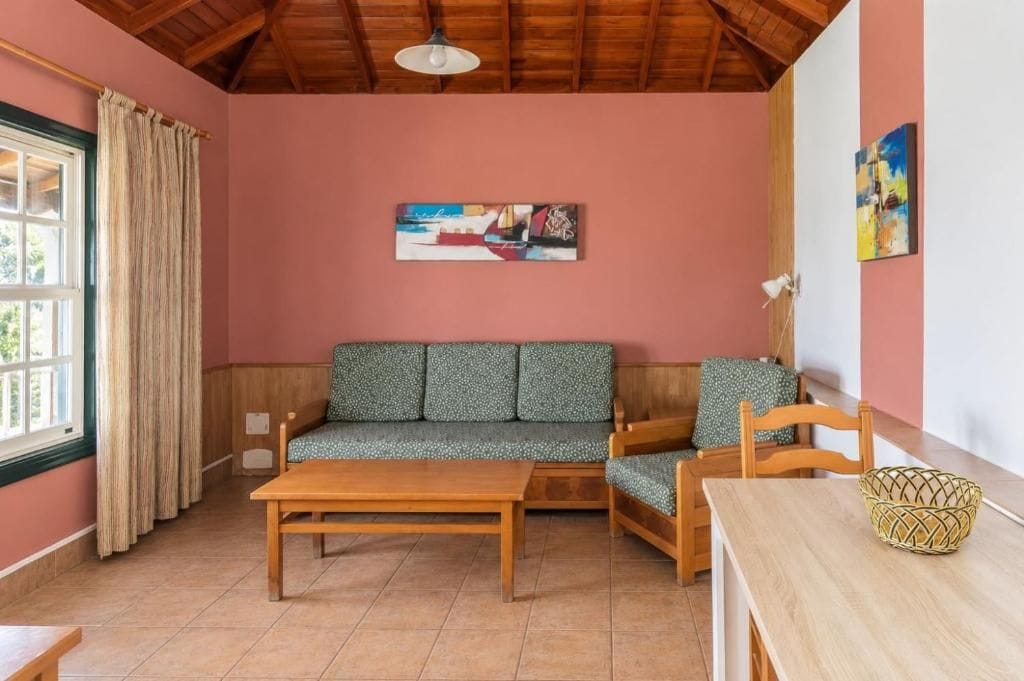 Quarto com teto de madeira, sofá com almofadas estampadas, mesa de madeira e janela com cortinas.