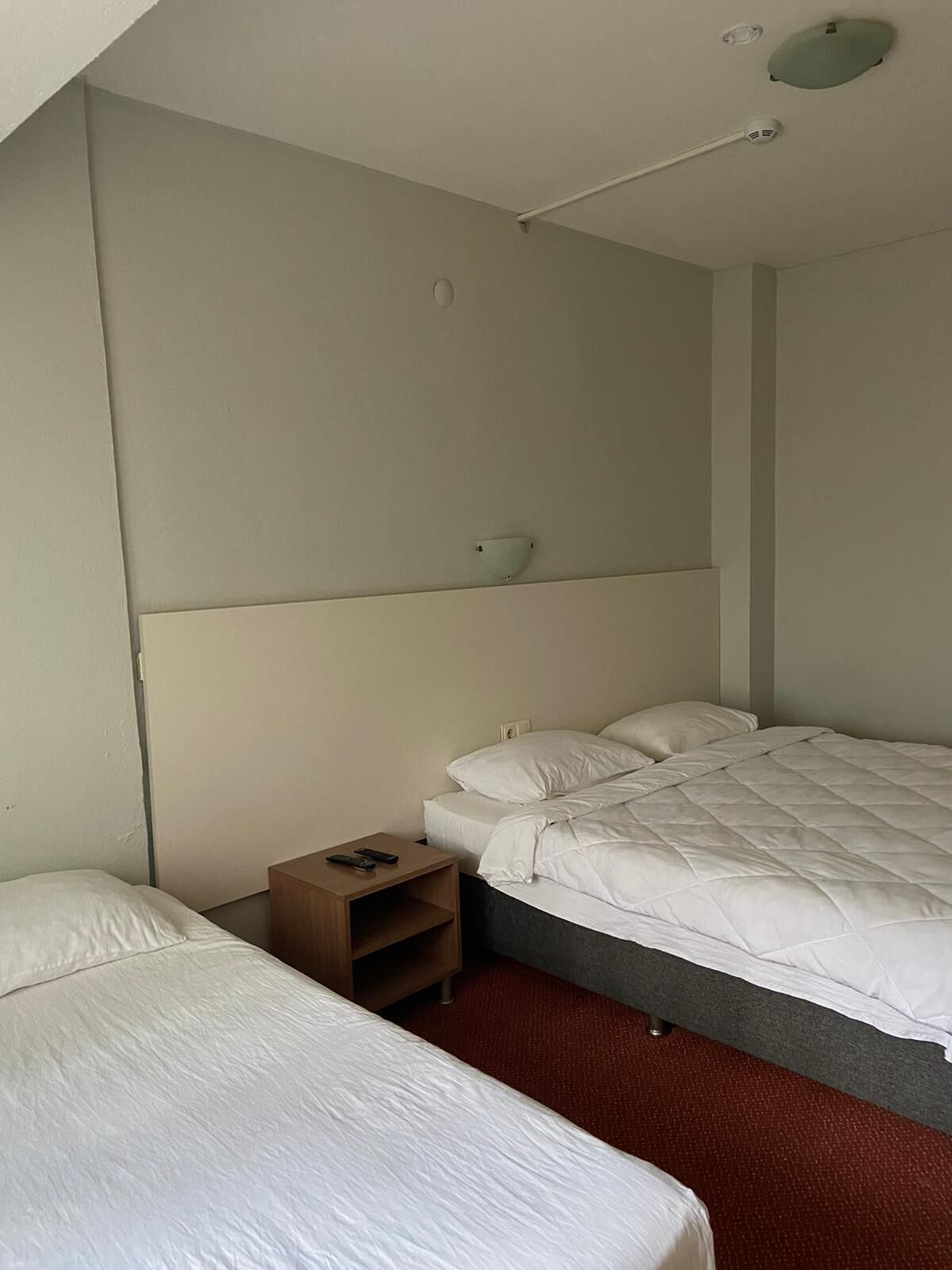 Quarto de hotel com duas camas de solteiro, uma pequena mesa de cabeceira de madeira, luminária fixada na parede e tapete vermelho.