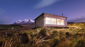 Uma casa moderna com grandes janelas, localizada numa colina gramada com uma montanha ao fundo.