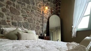Quarto com parede de pedra, cama com roupa de cama branca, mesa de cabeceira com abajur e janela com cortinas leves.